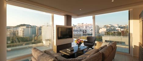 Sala de estar moderna com televisão grande, assentos confortáveis e vista para a cidade através de amplas janelas.