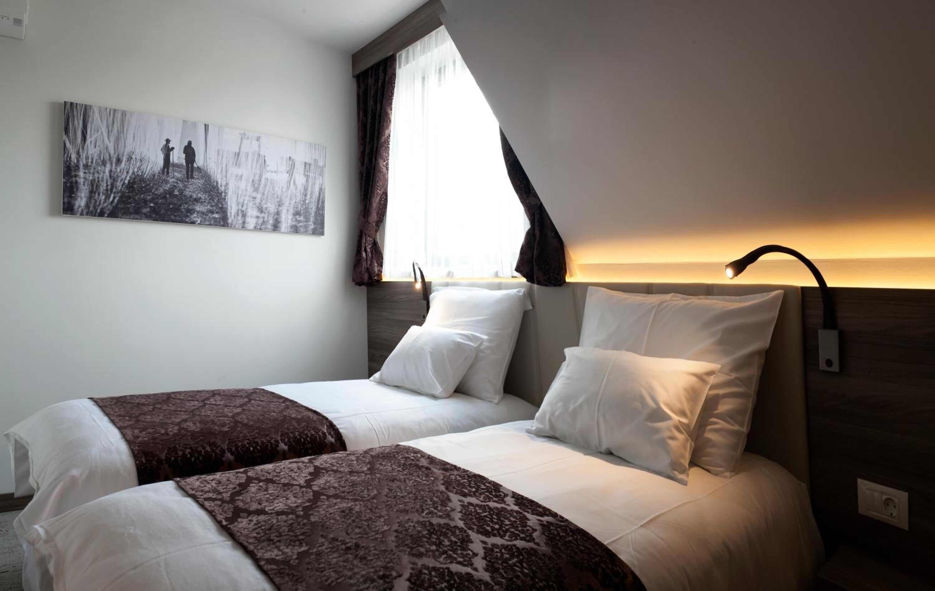Quarto de hotel com duas camas, uma janela grande com cortinas, um quadro na parede e uma mesa de cabeceira com abajur.