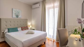 Quarto de hotel com uma cama grande, uma mesinha de cabeceira com abajur, uma cadeira, um vaso de planta e uma janela com cortinas.