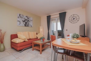 Uma sala de estar com um sofá vermelho, uma mesa de centro de vidro e uma mesa de jantar com cadeiras. Há um quadro na parede, um relógio e uma TV em um móvel.