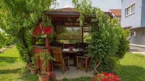 Uma pérgola de madeira com uma mesa e cadeiras, rodeada por plantas e flores em vasos.