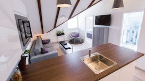 Cozinha moderna com bancada de madeira, pia de aço inoxidável, luminárias pendentes e vista para a sala de estar, com um sofá e uma poltrona.