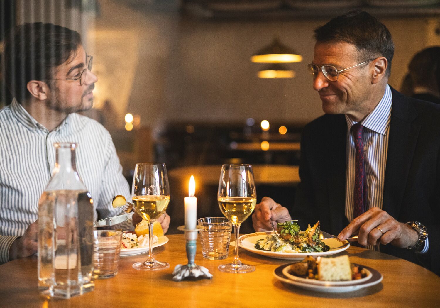Duas pessoas jantando em restaurante, com velas, copos de vinho e uma garrafa sobre a mesa.
