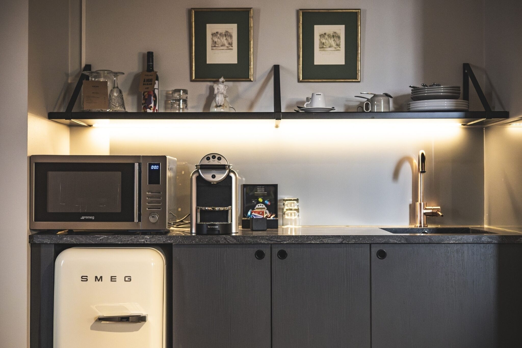 Uma cozinha com micro-ondas, cafeteira e frigorífico. Há quadros nas paredes acima do balcão.