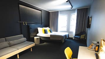 Quarto de hotel moderno com sofá, cama com almofadas, escrivaninha com cadeira e pequena mesa com garrafa e flores.