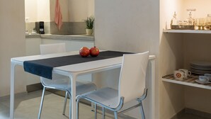 Uma área de refeições com uma mesa branca, duas cadeiras e uma planta em vaso. Dá para ver uma cozinha ao fundo, com uma pia e bancada.