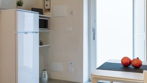 Uma cozinha compacta com geladeira, micro-ondas e uma pequena prateleira com uma planta em vaso. Há uma porta que dá para um quarto claro, com uma mesa e duas romãs sobre ela.