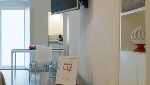 Uma sala de estar moderna com televisão de tela plana na parede, um letreiro de boas-vindas emoldurado, uma mesa de jantar com cadeiras e uma área de cozinha ao fundo.