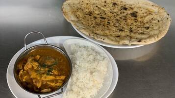 Uma refeição com uma tigela de curry, um prato de arroz branco e um pedaço de pão achatado sobre uma mesa de aço inoxidável.