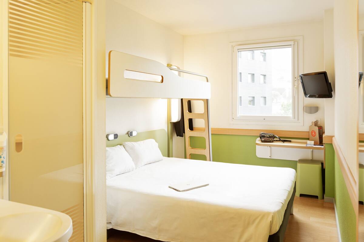 Quarto de hotel com uma cama beliche, uma escrivaninha com cadeira, uma TV fixada na parede e uma janela com vista para o exterior.
