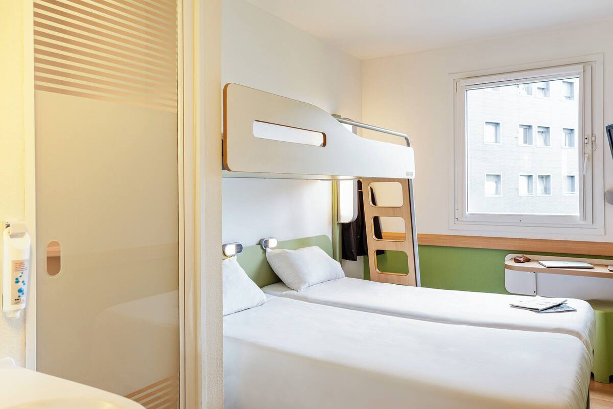 Quarto de hotel com duas camas, uma janela, uma escrivaninha e um banheiro.