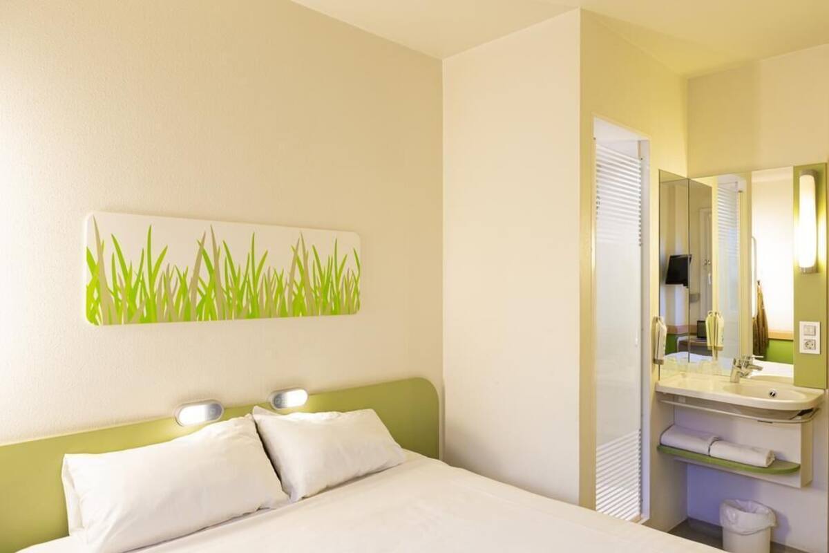 Quarto de hotel com cama, abajures, obra de arte na parede, banheiro com pia e espelho.