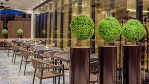 Uma área de refeições interna moderna, com mesas e cadeiras redondas, plantas esféricas decorativas e piso estampado.