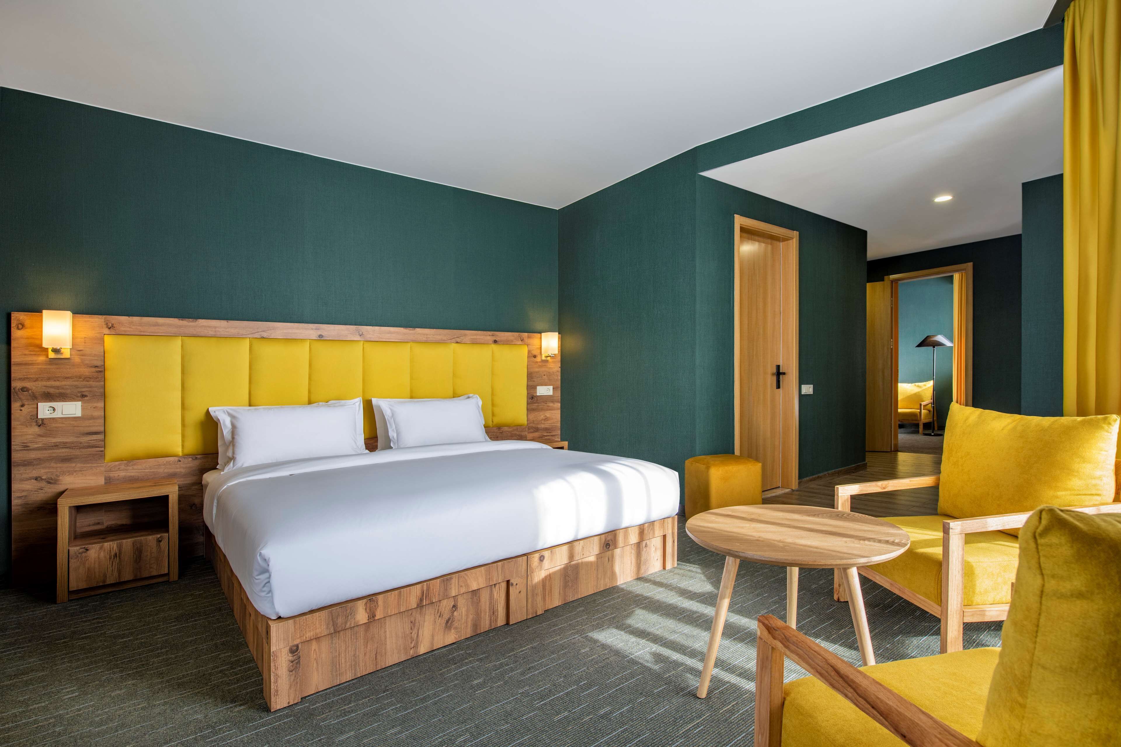 Quarto de hotel com cama, cabeceira de madeira, parede de destaque amarela, mesa de cabeceira de madeira pequena, poltrona amarela, mesa lateral de madeira e porta que dá para outro quarto.