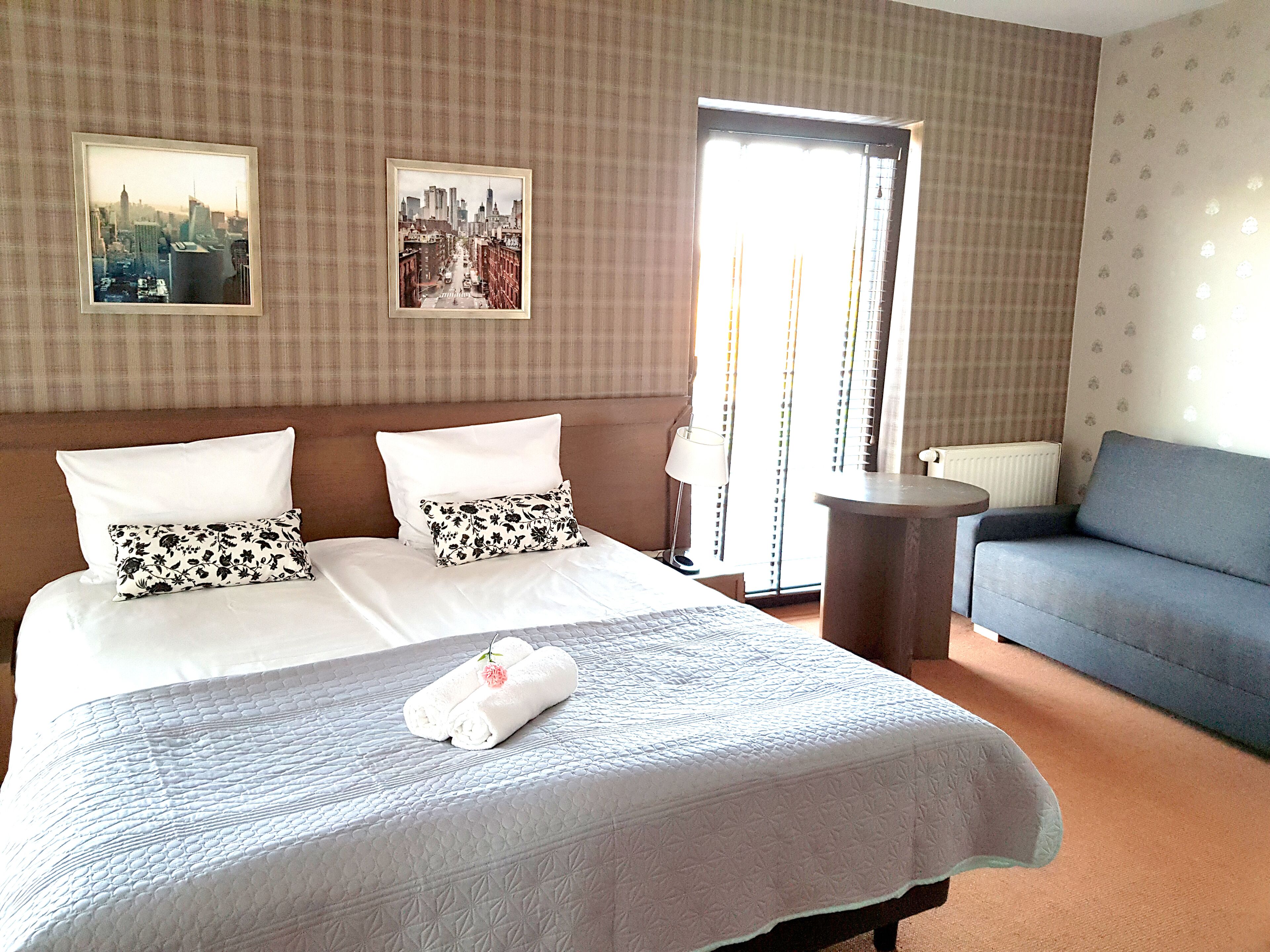 Quarto de hotel com uma cama grande, duas almofadas, um sofá azul e duas imagens emolduradas com vista para a cidade.