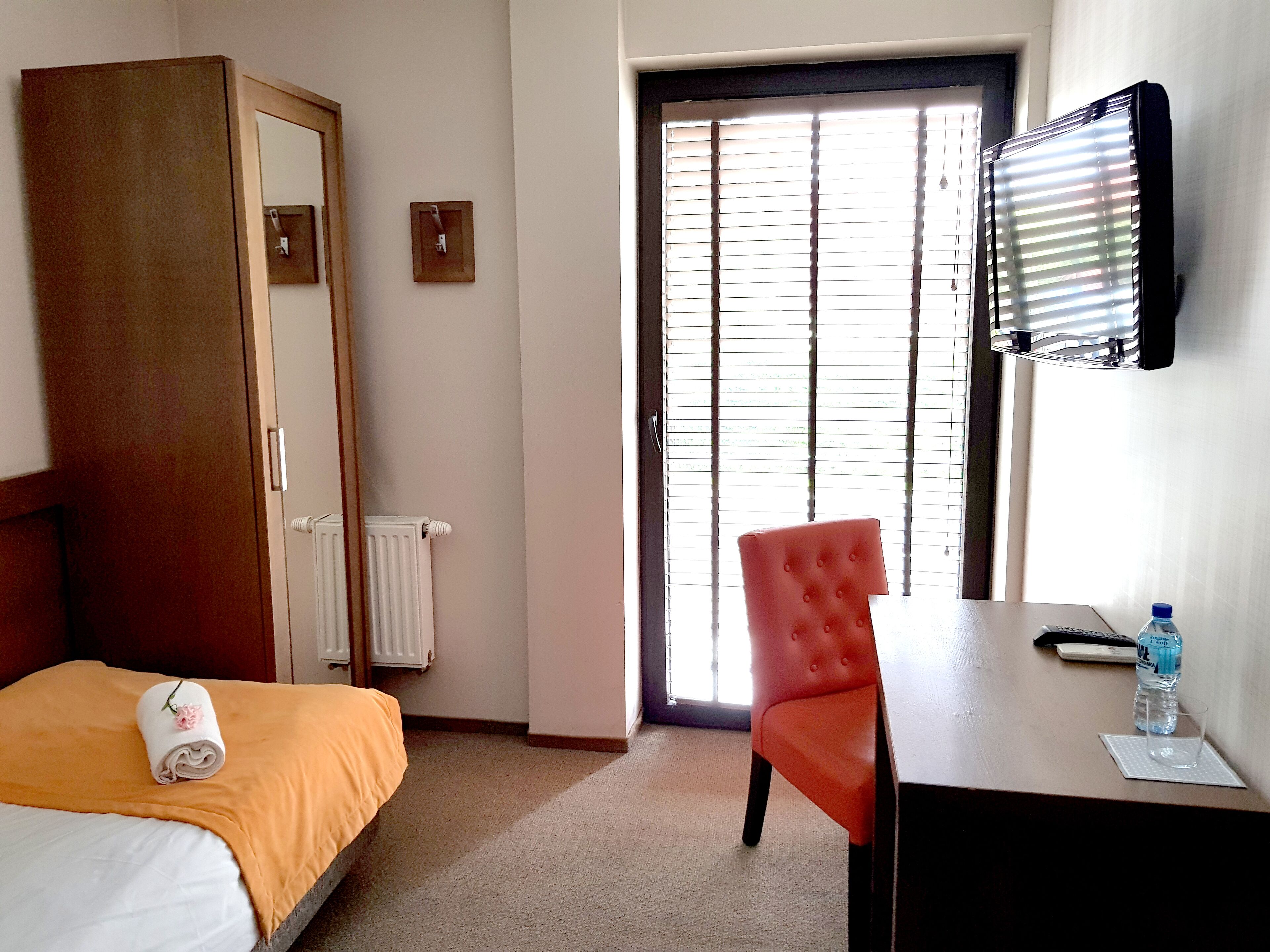 Quarto de hotel com cama, escrivaninha, cadeira, radiador, janela com persianas e televisão fixada na parede.