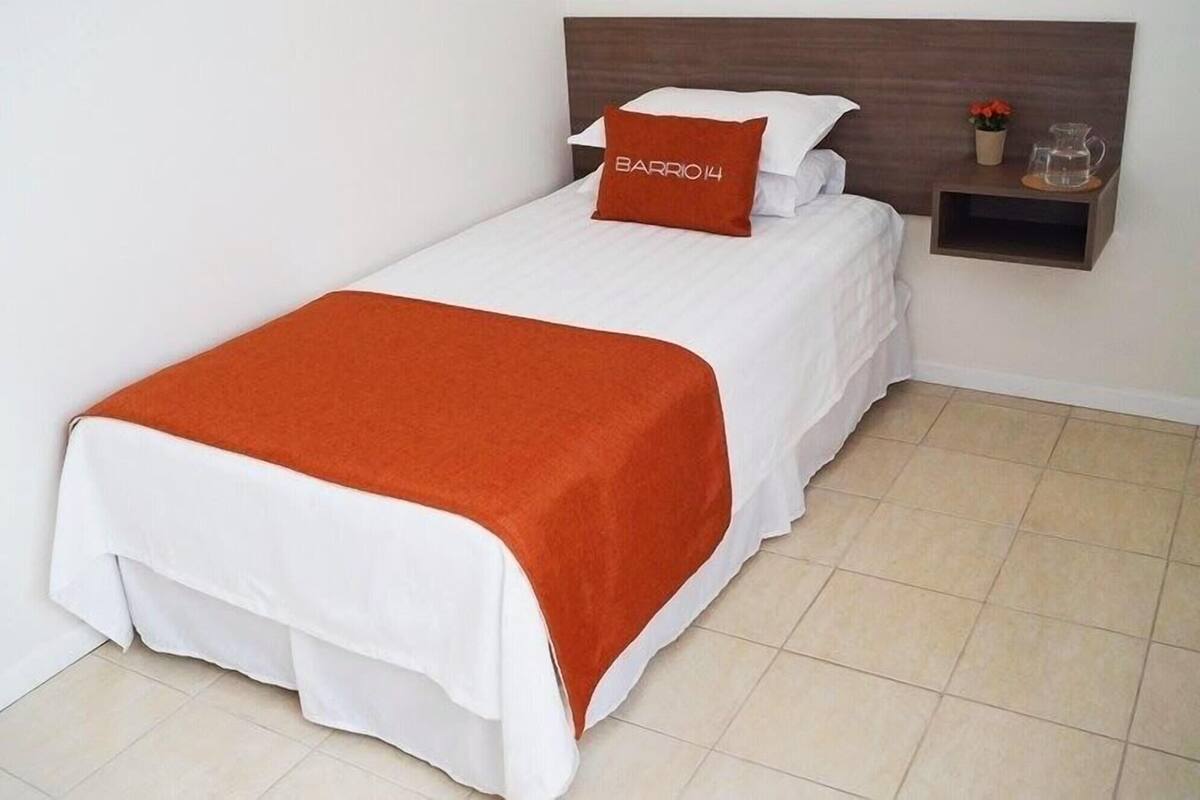 Uma cama de solteiro com roupa de cama branca e um cobertor vermelho, cabeceira de madeira, mesa de cabeceira pequena com um vaso de flores, e chão revestido com azulejos.