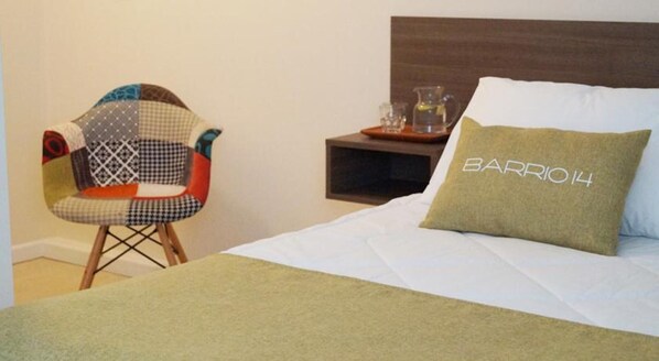 Quarto de hotel com cama, uma cadeira colorida, mesa de cabeceira com um copo de água e um travesseiro com o texto "BARRIO14".