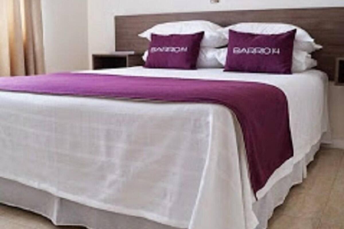 Uma cama bem arrumada com edredom branco e roxo, duas almofadas com a inscrição "BAGHDAD" e cabeceira de madeira.
