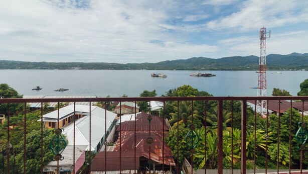 Vista costeira com barcos na água, uma torre de comunicação vermelha e edifícios com telhados vermelhos.