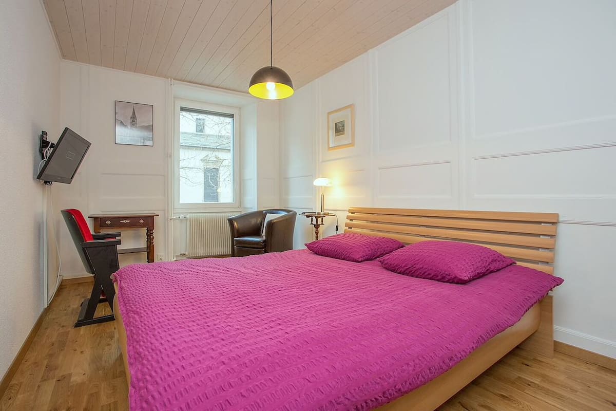 Quarto com cama de madeira, escrivaninha com cadeira, televisão na parede e janela com vista para edifícios.