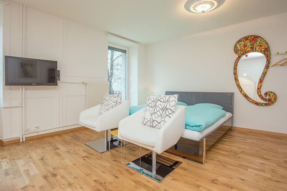 Quarto moderno com uma cama, duas poltronas brancas, uma TV e um espelho grande.