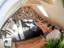 Quarto estilo loft com teto curvo, cama com edredom preto e branco, piso de madeira e claraboia.