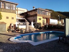 Uma área de piscina com um terraço de pedra, mobiliário de exterior e um edifício com varanda e escadas.