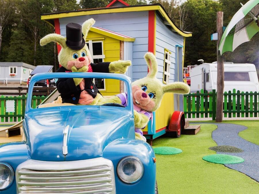 Um carro azul antigo com duas pessoas em um cenário divertido, uma casa pequena e uma cerca verde.