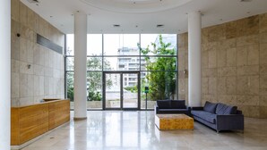 Um hall amplo com balcão de recepção, um sofá e uma mesa de centro. Grandes janelas oferecem vista para o exterior.