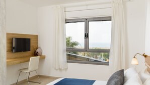 Quarto com cama, televisão em prateleira de madeira, escrivaninha com cadeira e janela com cortinas.
