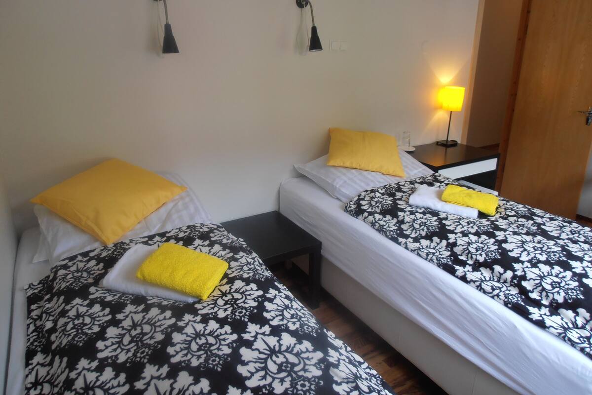 Quarto de hotel com duas camas de solteiro, cada uma com colcha estampada com flores e travesseiro amarelo. Há luminárias fixadas na parede e mesas de cabeceira com abajures.