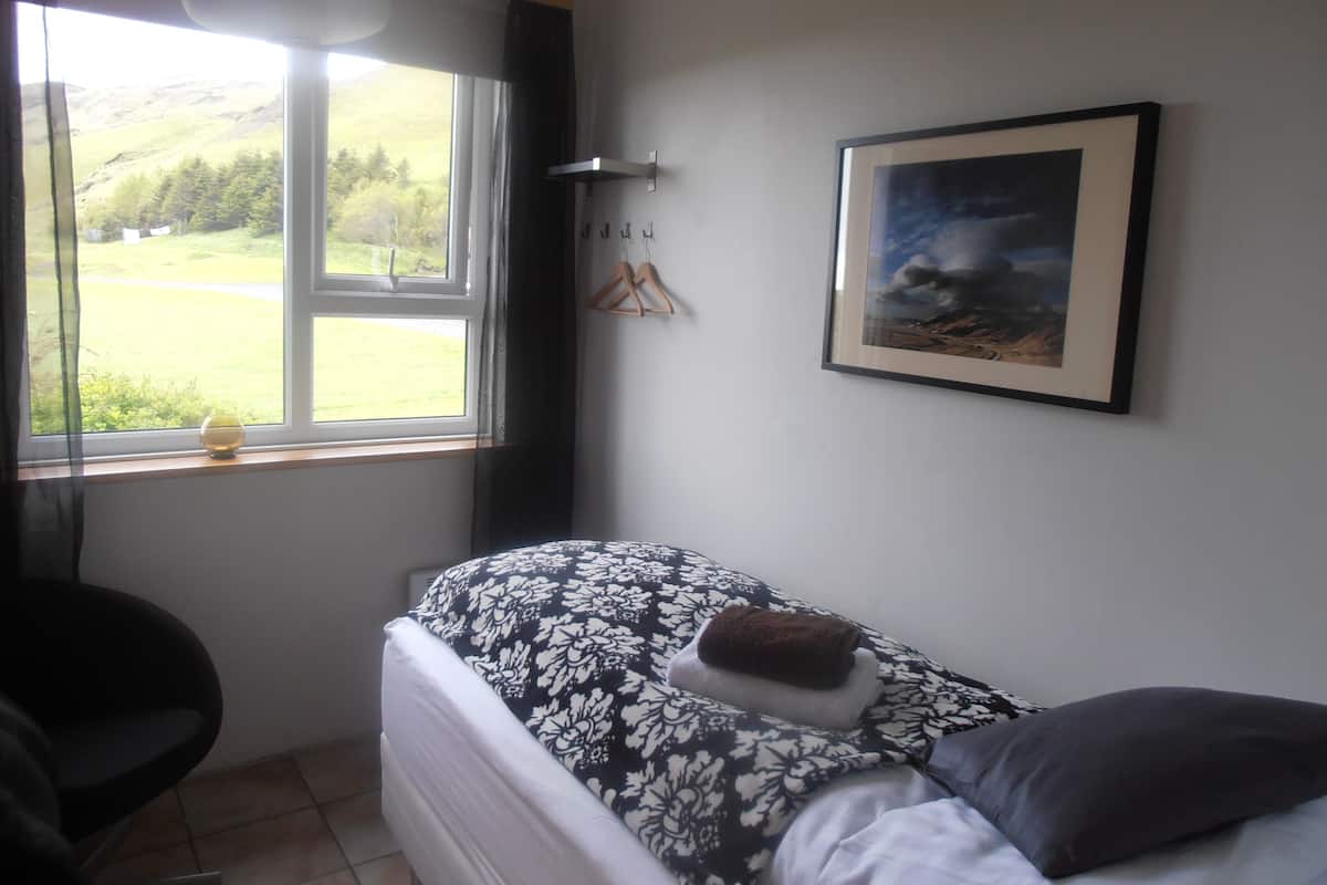 Um quarto com uma cama, uma janela com vista para uma paisagem verde, um lustre pendurado e um quadro na parede.