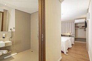 Quarto de hotel com chuveiro, pia com artigos de higiene e cama com colcha branca, além de TV fixada na parede.