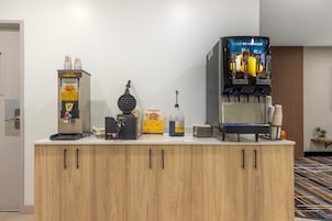 Uma cozinha compacta com máquina de waffle, dosador de massa para waffle, garrafa de calda para waffle e um distribuidor de bebidas geladas.