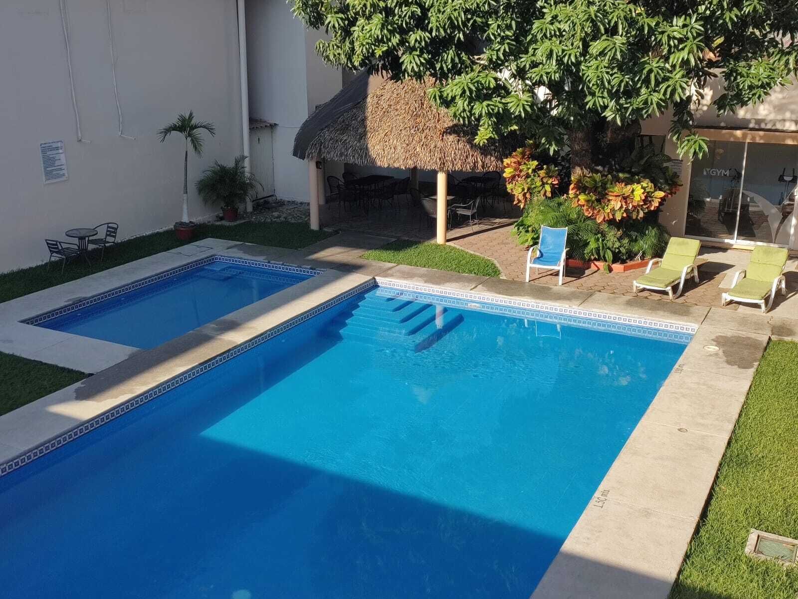 Piscina com duas áreas distintas, rodeada por grama e plantas em vasos. Há espreguiçadeiras e uma estrutura com telhado de palha por perto.
