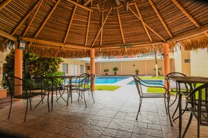 Uma área de estar externa com telhado de palha, piscina e plantas em vasos.