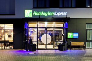 Exterior de um hotel Holiday Inn Express, com uma grande placa iluminada, entrada de vidro e um banco na área externa.
