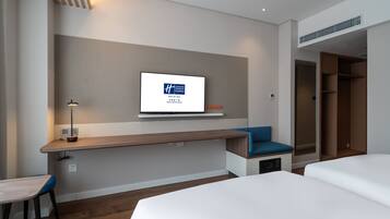 Quarto de hotel com cama, escrivaninha com luminária, televisão na parede e armário.
