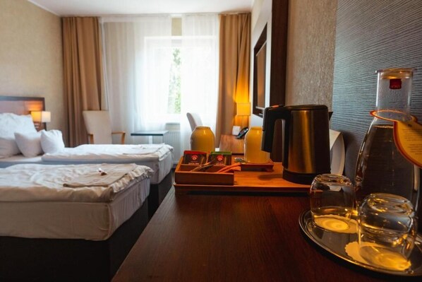 Quarto de hotel com duas camas, uma mesa de madeira com um tabuleiro de copos e uma chaleira, e uma janela grande com cortinas.