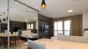 Quarto de hotel moderno com área de refeições, uma grande televisão de tela plana, um sofá e uma janela ampla com cortinas.