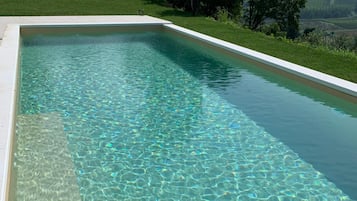 Piscina retangular com água cristalina, rodeada por um deck de concreto e uma área gramada com árvores ao fundo.