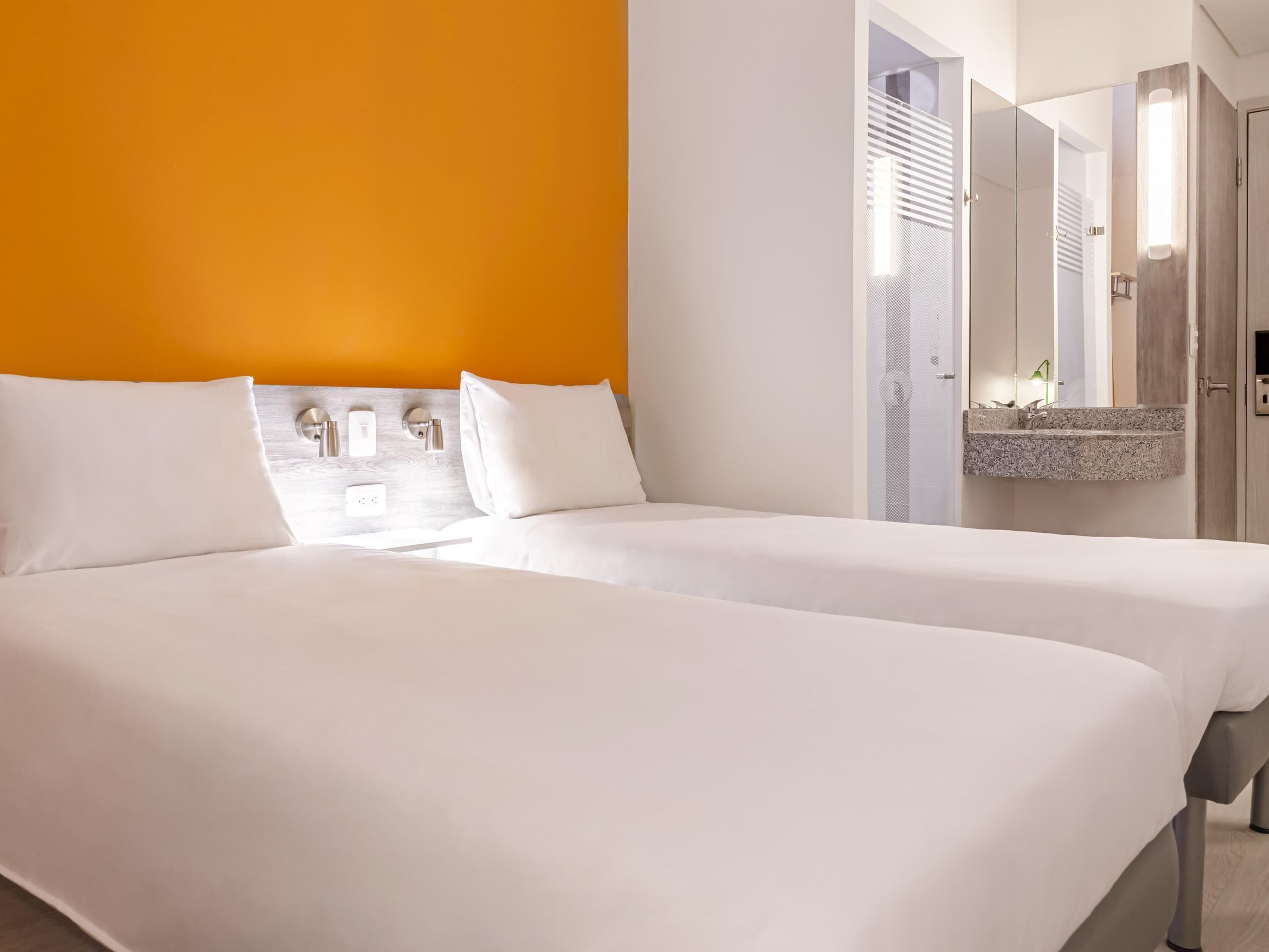 Quarto de hotel com duas camas, uma parede de destaque laranja vibrante, um espelho grande e uma bancada de granito.
