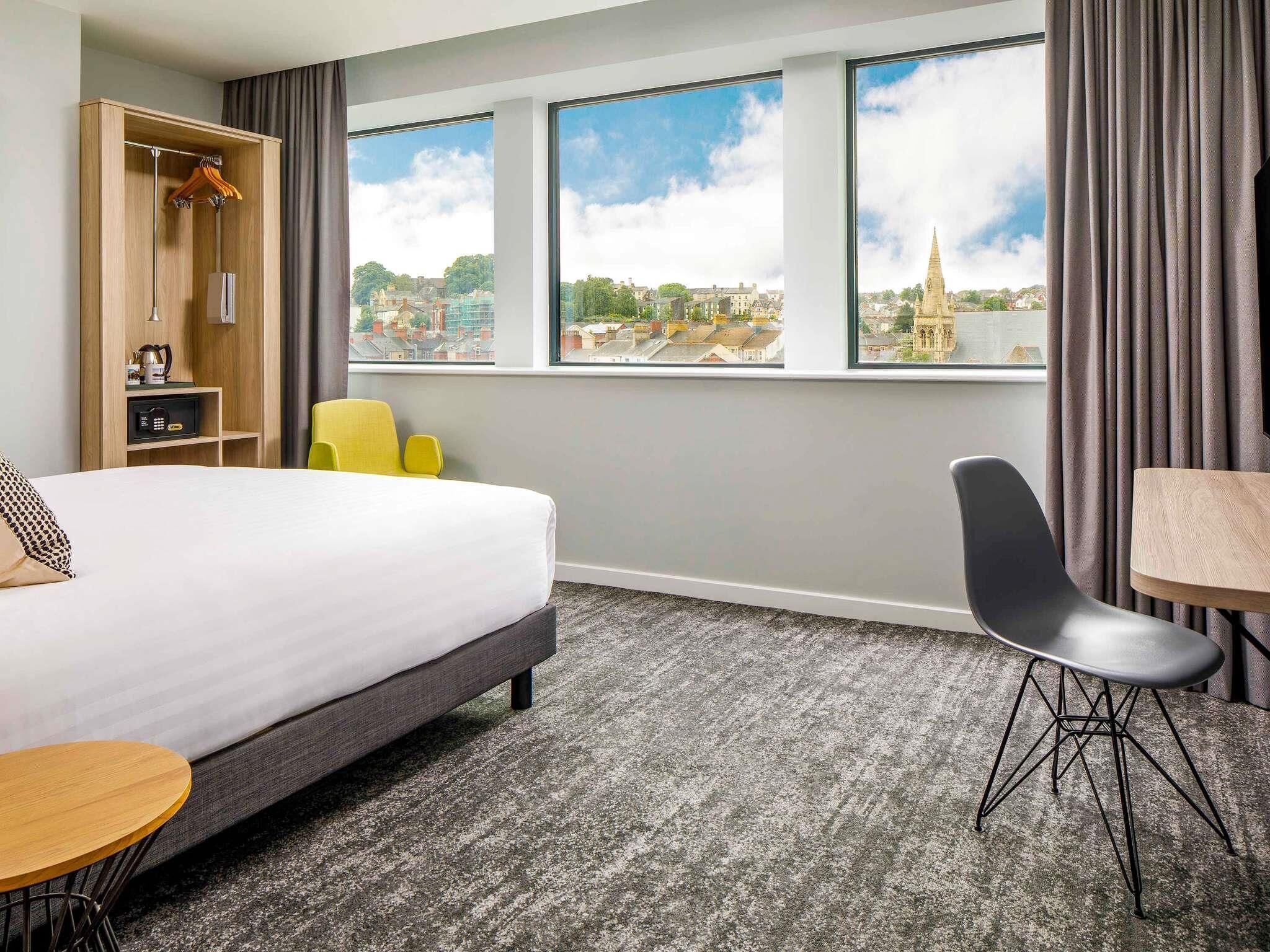 Quarto de hotel com cama, cadeira, mesa pequena, guarda-roupa e vista para a paisagem urbana através de janelas amplas.