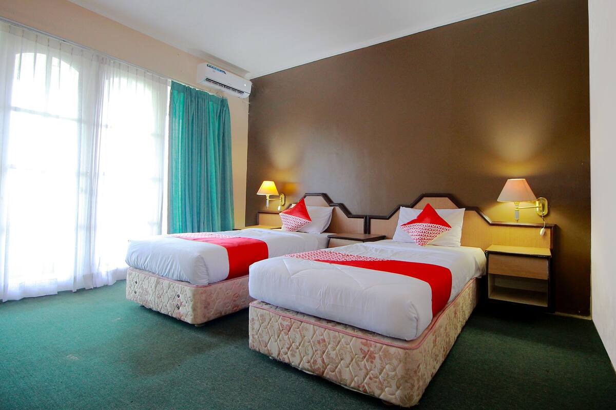 Quarto de hotel com duas camas, uma mesa de cabeceira com luminárias, uma janela com cortinas e um aparelho de ar condicionado.