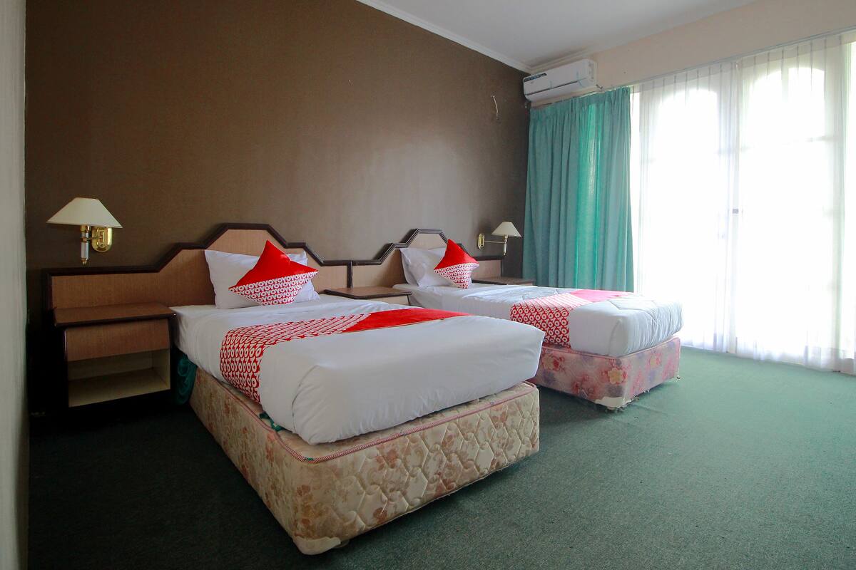 Quarto de hotel com duas camas, cada uma com travesseiro e cobertor. Há duas mesas de cabeceira com luminárias, uma janela com cortinas e um ar-condicionado na parede.