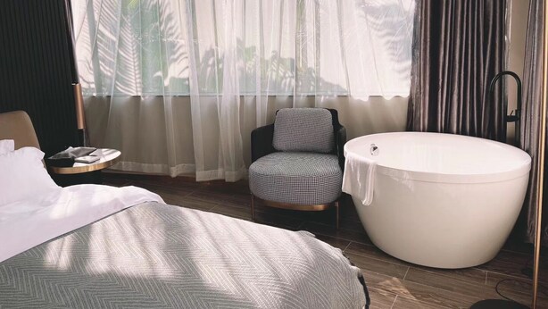 Quarto de hotel com uma cama grande, uma banheira redonda, uma poltrona pequena, uma mesinha de cabeceira e cortinas translúcidas que deixam entrar luz natural.