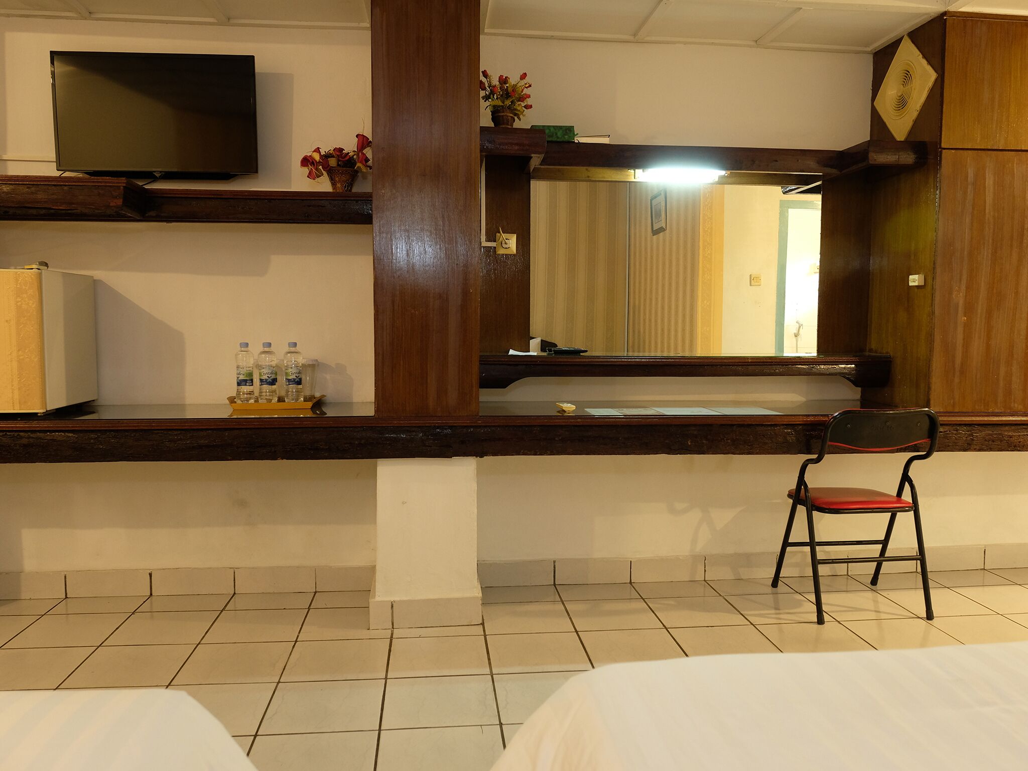 Quarto de hotel com uma cama, uma cadeira, uma televisão, um espelho, uma prateleira com garrafas de água e uma escrivaninha de madeira.