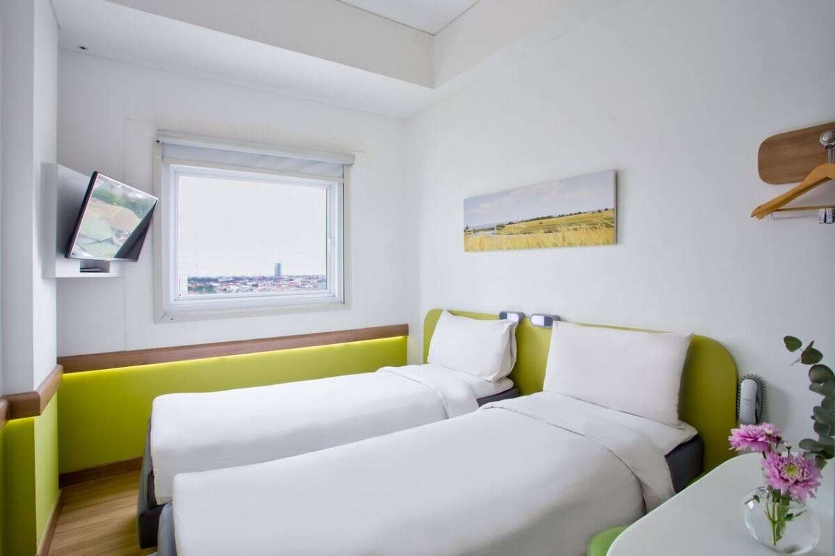 Quarto de hotel com duas camas, televisão, janela com vista para a cidade, um quadro e uma mesinha com flores.