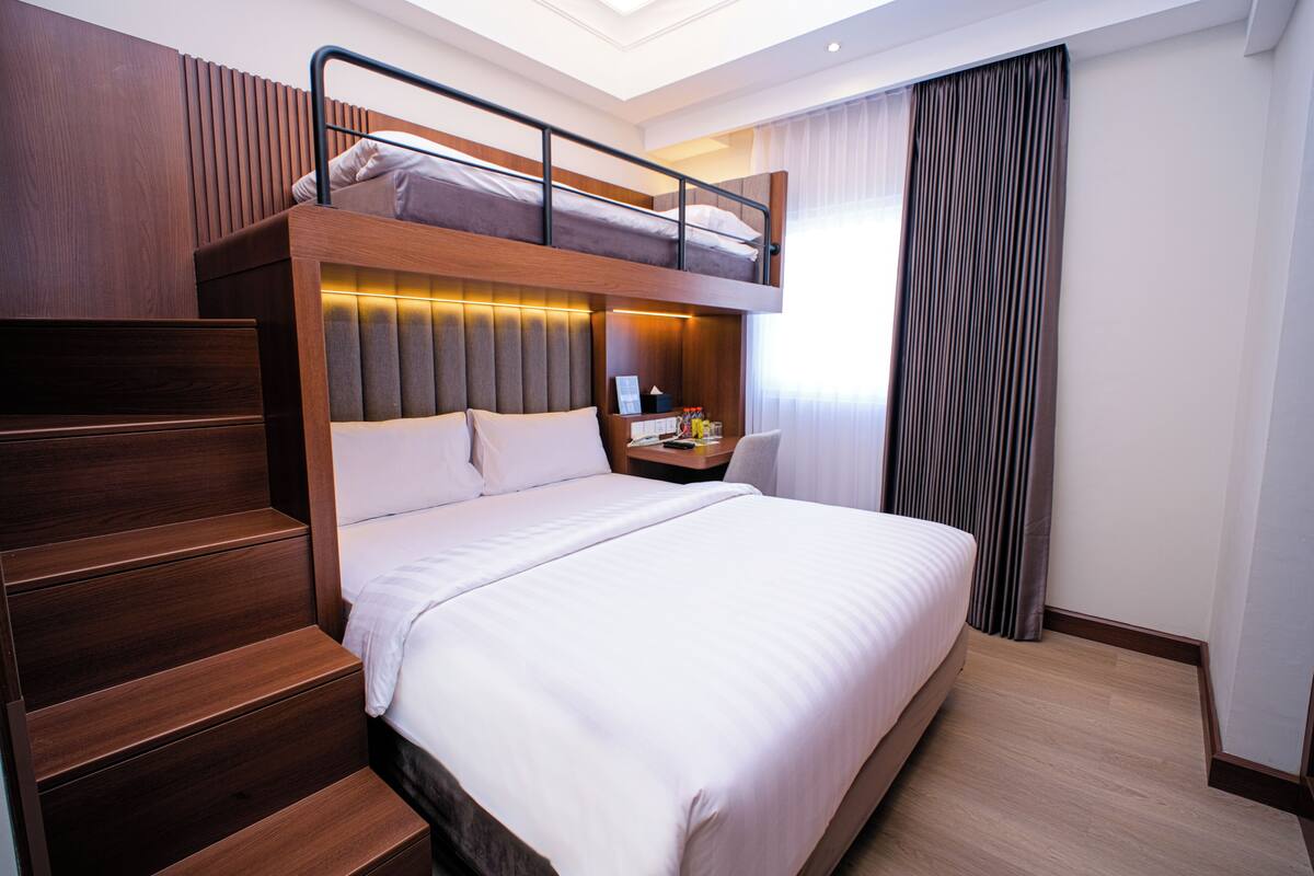 Quarto de hotel com beliche, escrivaninha com cadeira e uma janela grande com cortinas.